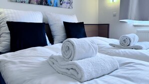 Quarto de hotel com cama bem arrumada, almofadas brancas e azul escuras, e toalha branca enrolada sobre a cama.