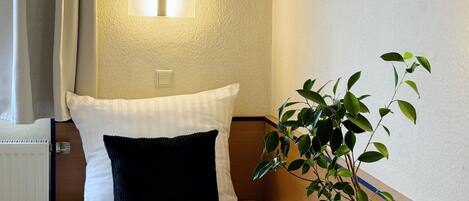 Quarto de hotel com cama, mesa de cabeceira, planta em vaso e luminária na parede.