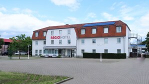 Edifício REGIOHOTEL com telhado de telhas vermelhas, paredes externas brancas e área de entrada pavimentada.