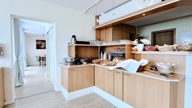 Cozinha moderna com armários de madeira, bancada com diversos objetos e área de refeições visível através de uma porta aberta.
