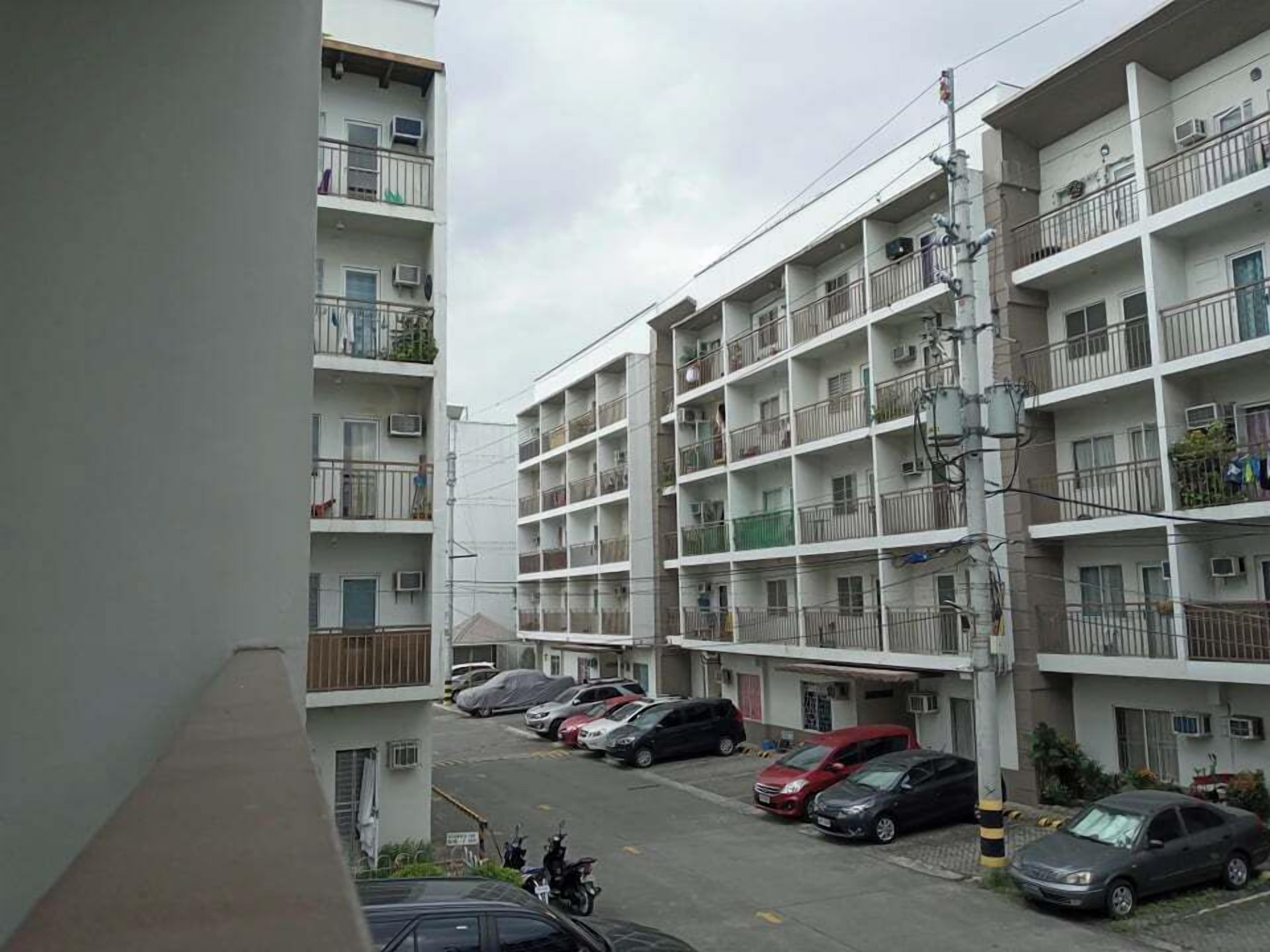 Edifício residencial de vários andares, com varandas, área de estacionamento com vários carros e unidades de ar condicionado visíveis.
