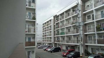 Edifício residencial de vários andares, com varandas, área de estacionamento com vários carros e unidades de ar condicionado visíveis.