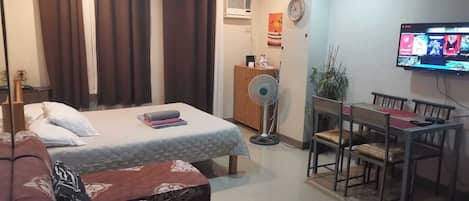 Quarto de hotel com cama, sofá, mesa de jantar com cadeiras, televisão e ventoinha.