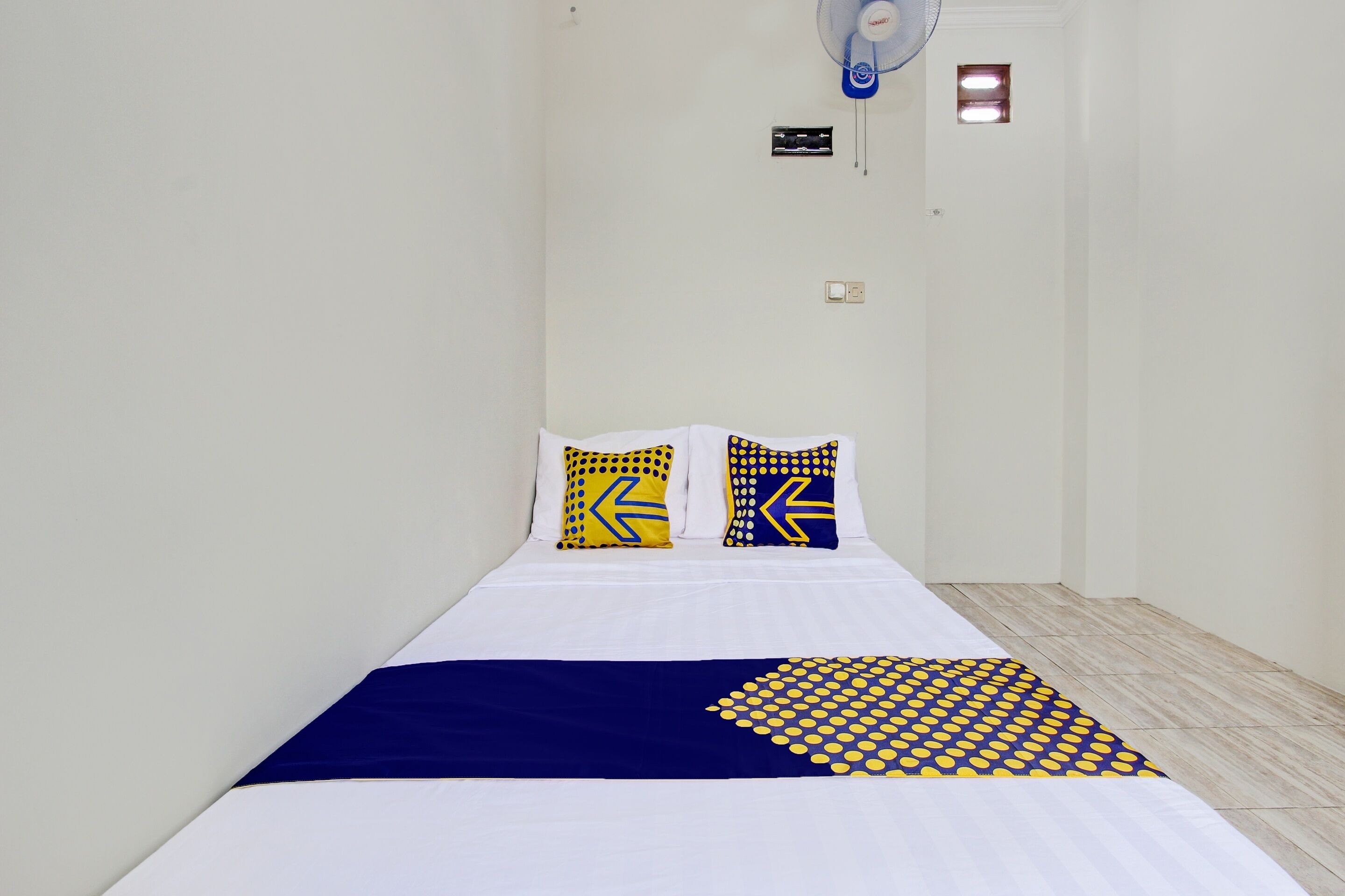 Quarto de hotel com uma cama, duas almofadas com padrões geométricos, um ventilador fixado na parede e uma luminária pequena.