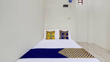 Quarto de hotel com uma cama, duas almofadas com padrões geométricos, um ventilador fixado na parede e uma luminária pequena.