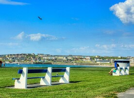 Uma área gramada com um banco azul e branco, um pequeno abrigo e vista para uma cidade costeira com edifícios e uma praia.