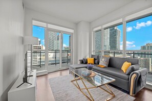 Uma sala de estar moderna com um sofá cinzento, almofadas amarelas, uma mesa de centro de vidro e vista para edifícios altos através de grandes janelas.
