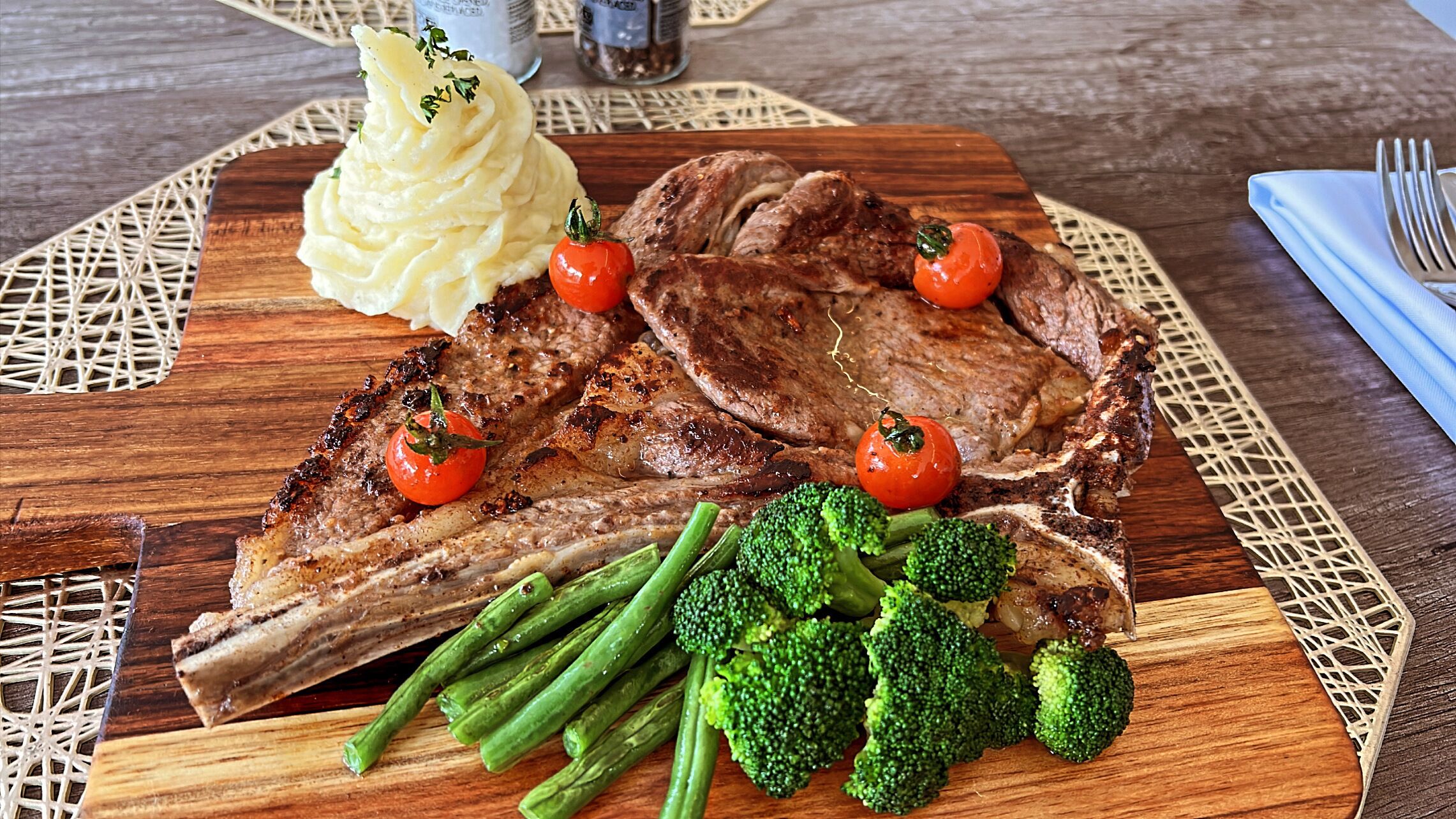 Uma mesa de madeira com uma refeição de bife grelhado, batatas assadas, tomates cereja e brócolis.