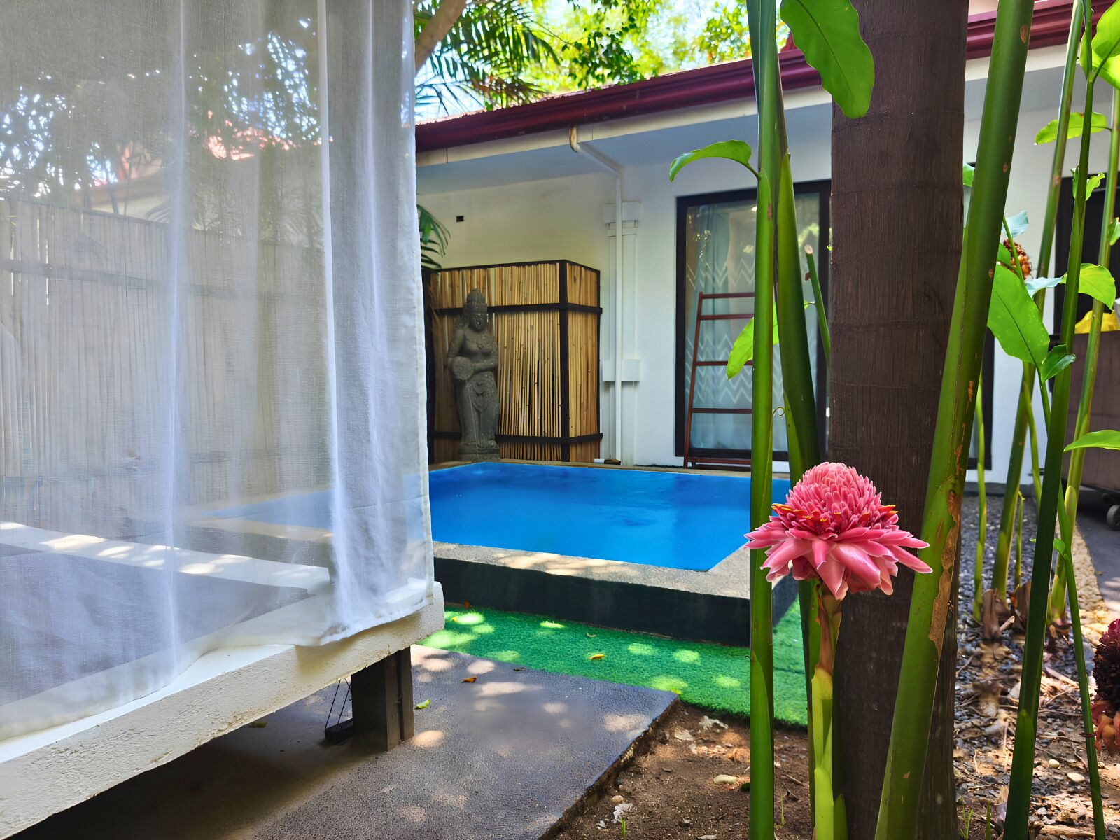 Área de piscina com um espaço de estar coberto, uma estátua e uma flor rosa.