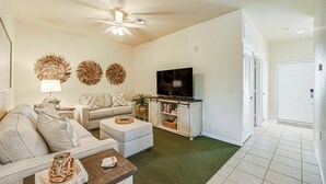 Uma sala de estar com sofá, apoio para os pés, mesa de centro, televisão e peças decorativas na parede.