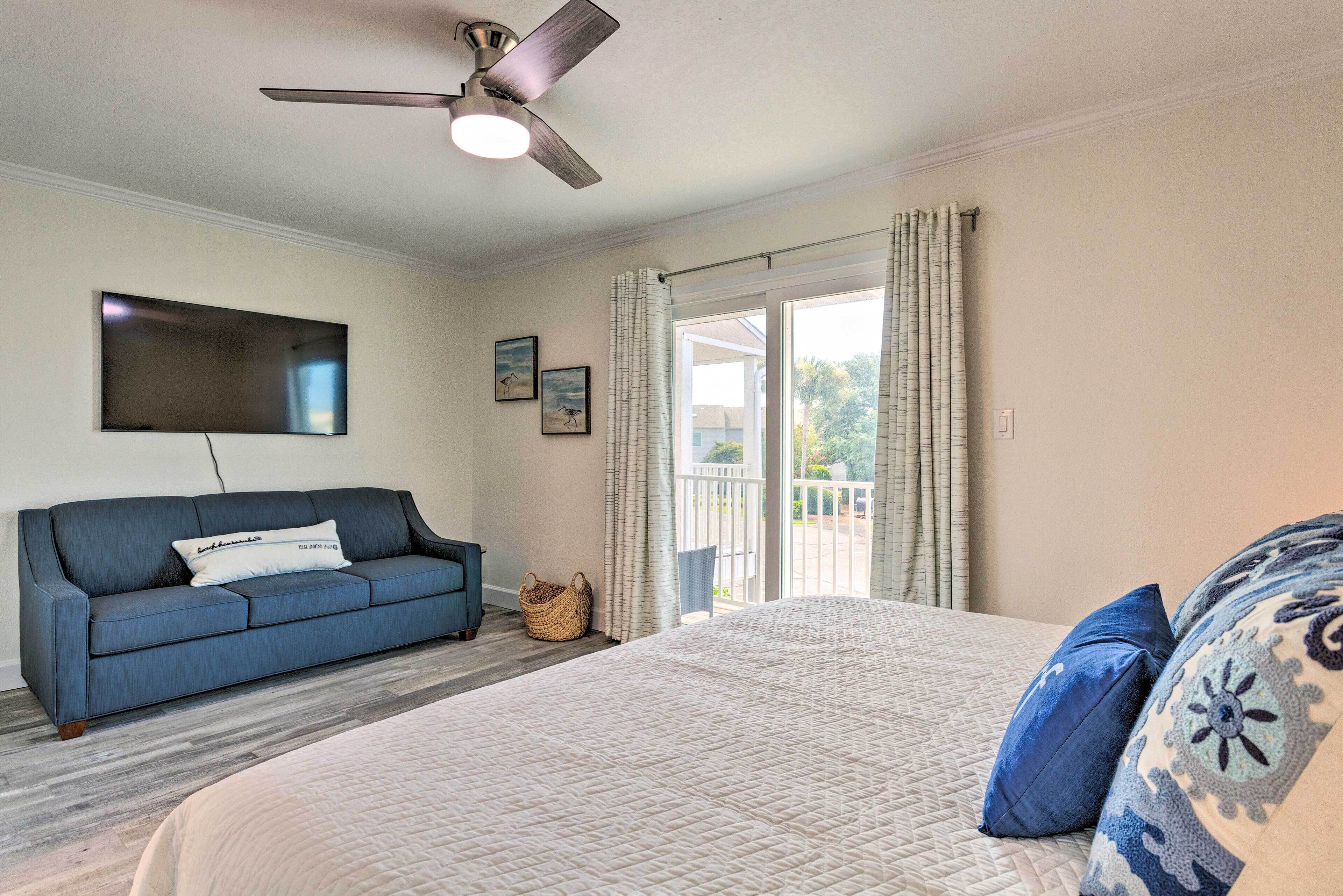 Quarto com cama, sofá azul, ventilador de teto, televisão de tela plana e varanda com cortinas.