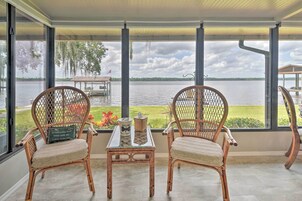 Uma área de estar coberta ao ar livre, com cadeiras de vime e uma mesa, com vista para um lago com um píer e uma área gramada.