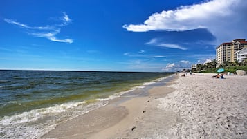 Uma praia com céu claro, mar calmo e edifícios ao fundo.