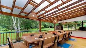 Uma área de refeições ao ar livre coberta, com uma longa mesa de madeira e bancos, cercada por uma pérgola de madeira com luzes penduradas.