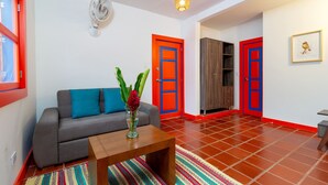 Uma sala de estar com sofá, mesa de centro, ventilador e porta com detalhes em vermelho e azul.