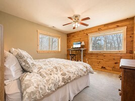 Quarto com cama, escrivaninha, computador, duas janelas e ventoinha no teto.