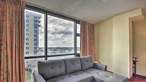 Sala moderna com sofá seccional cinza, grandes janelas com cortinas e vista para o horizonte urbano.