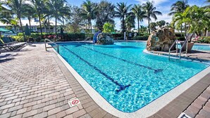 Piscina azul cristalina com um elemento de pedra, rodeada por palmeiras e espreguiçadeiras.