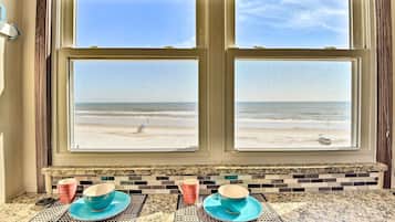 Uma cozinha com vista para a praia através de uma janela, uma bancada com um jogo de toalhas de mesa, e duas tigelas em cima.
