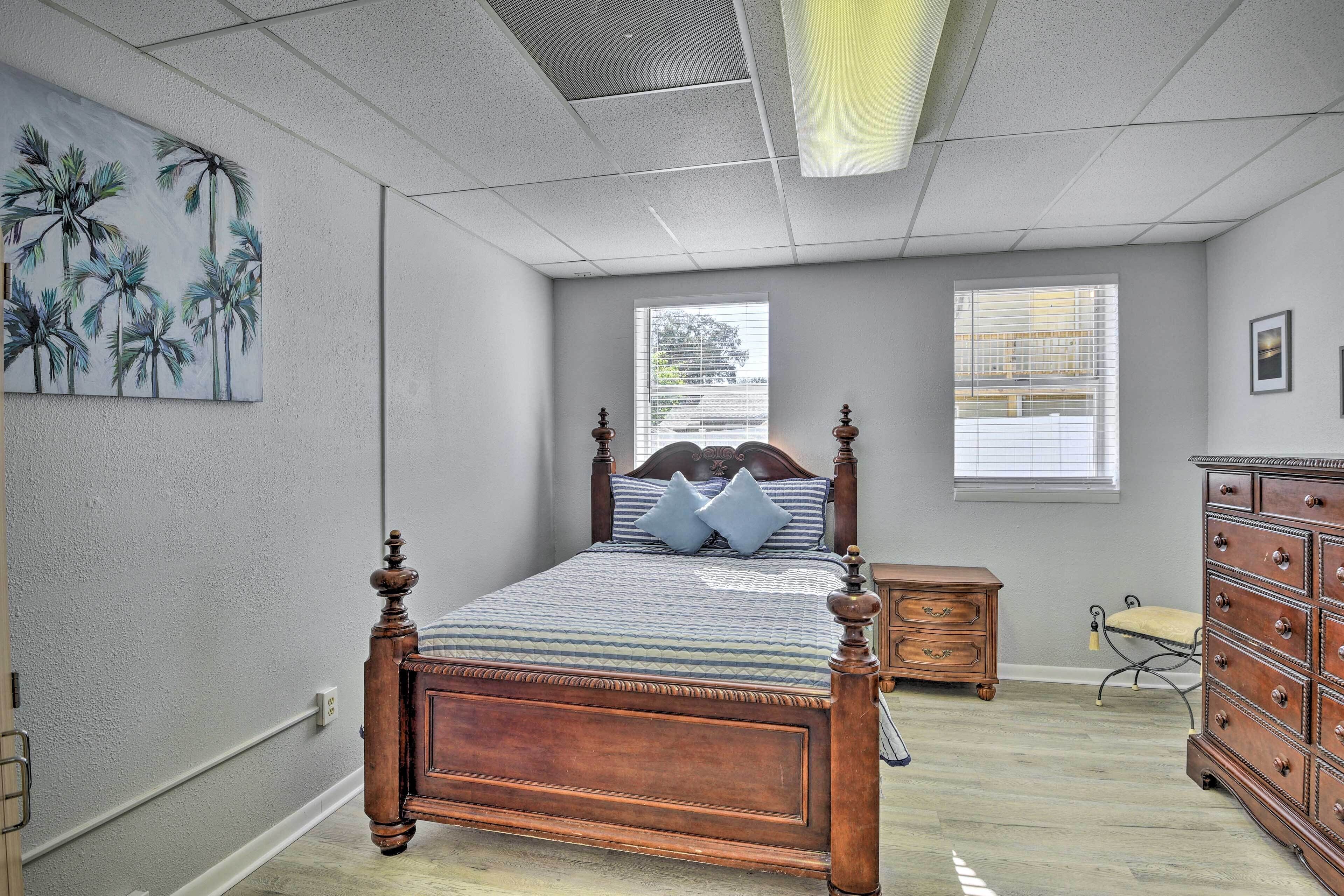 Quarto com cama de madeira, duas janelas, cômoda e mesa de cabeceira.