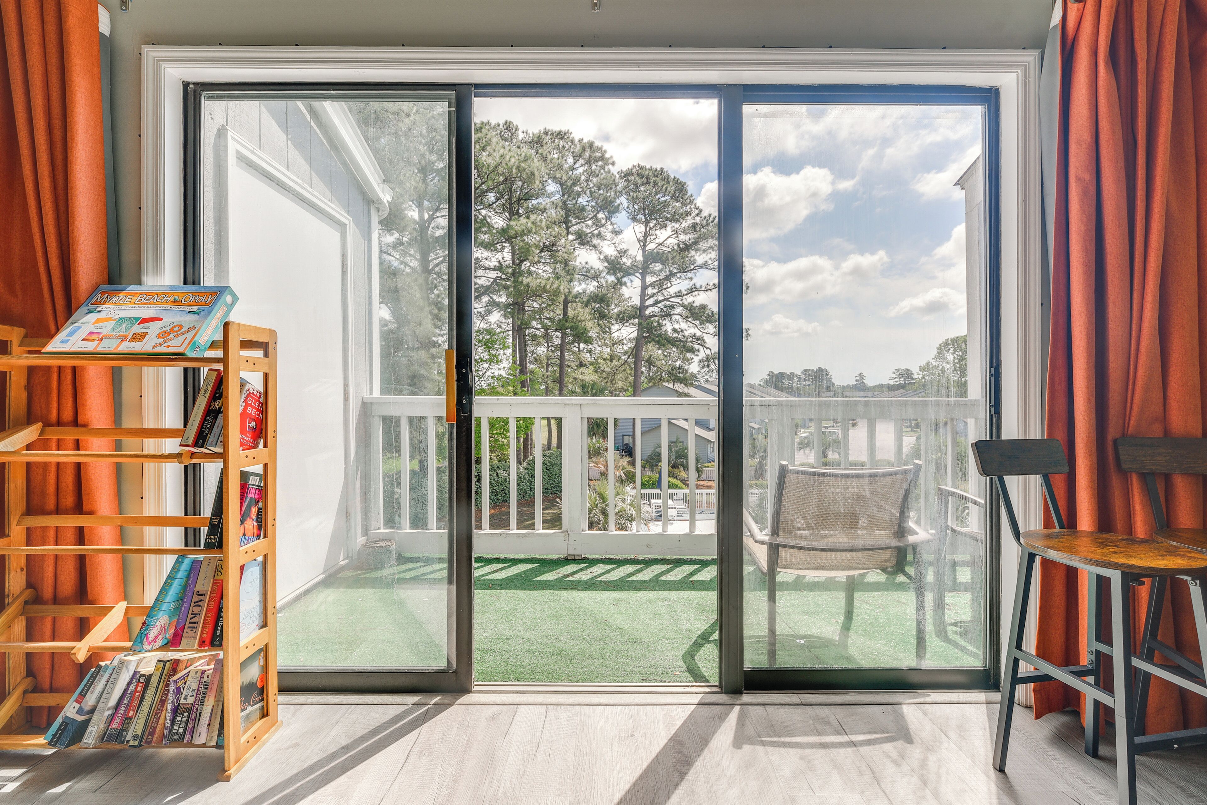 Quarto com porta de vidro deslizante para varanda com vista para árvores e cerca branca. Há uma estante de madeira com livros e uma cadeira.