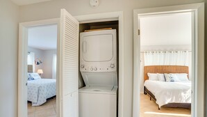 Quarto de hotel compacto com máquina de lavar, uma cama com roupa de cama branca e uma mesa de cabeceira com abajur.
