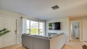 Uma sala de estar moderna com um sofá seccional cinza, uma TV de tela plana na parede e uma grande janela que oferece iluminação natural.
