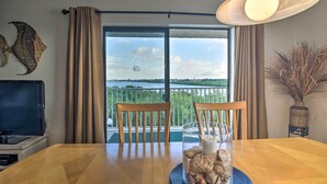 Uma sala de jantar com mesa, cadeiras, uma TV e vista para a água através de grandes janelas.