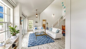 Uma sala de estar espaçosa com um sofá branco, uma mesa de centro de vidro e um tapete azul. Há uma escada com corrimão de madeira que leva a outro andar.