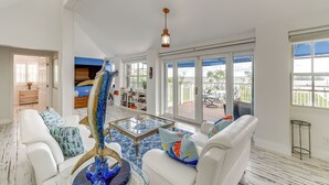 Uma sala de estar espaçosa com móveis brancos, uma mesa de centro de vidro e vista para o exterior por grandes janelas.