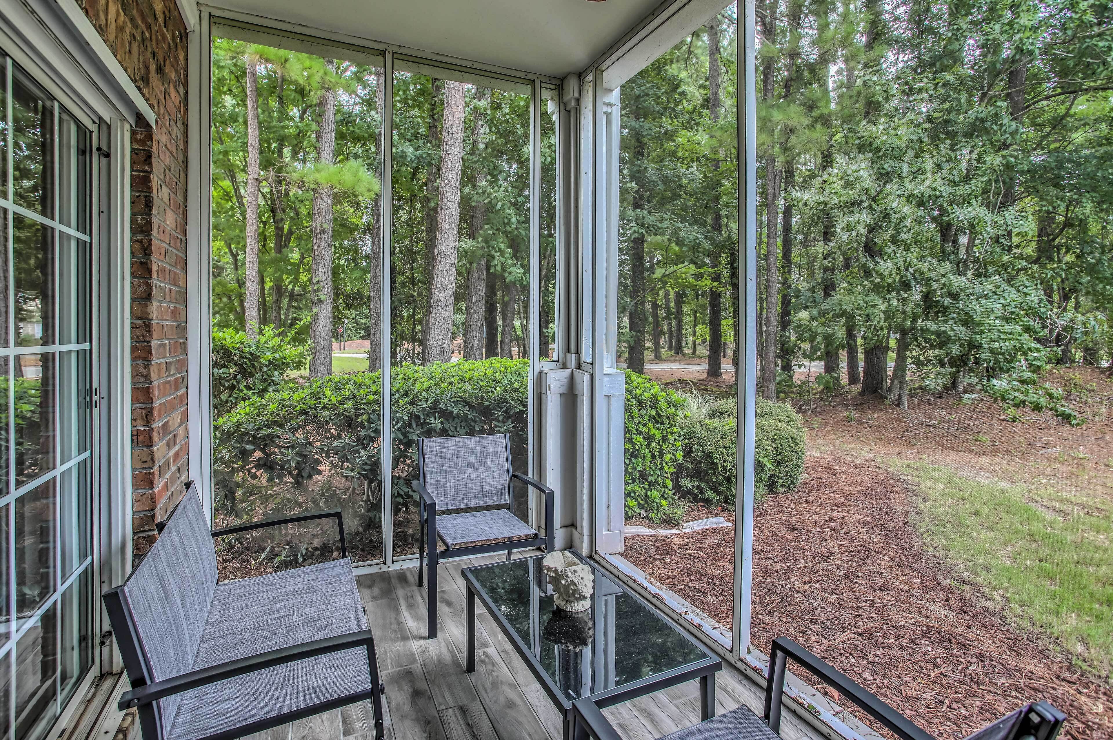 Uma área externa coberta com duas cadeiras e uma mesa, cercada por paredes de vidro com vista para uma área arborizada.