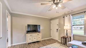 Quarto com ventilador de teto, televisão em uma cômoda, janela com cortinas e tapete estampado.