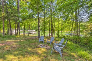 Um camping com três cadeiras Adirondack, um braseiro e vista para um lago rodeado por árvores.