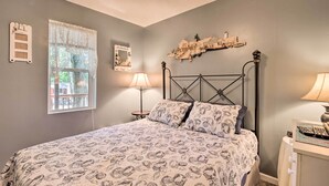 Quarto com cama, abajures, mesa de cabeceira, janela com cortinas de renda e prateleira na parede com objetos decorativos.
