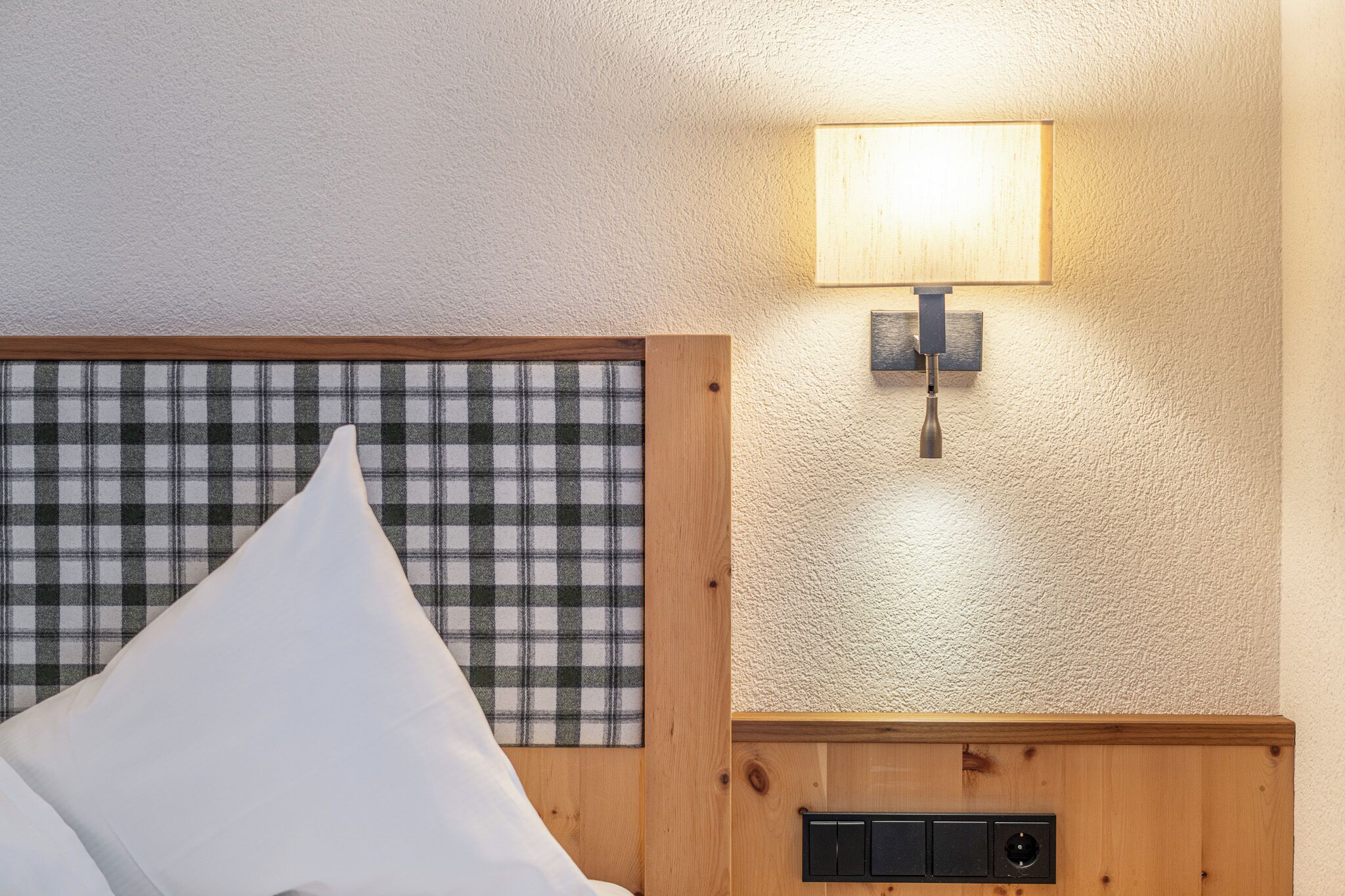 Quarto de hotel com cabeceira, luminária fixada na parede e tomada.
