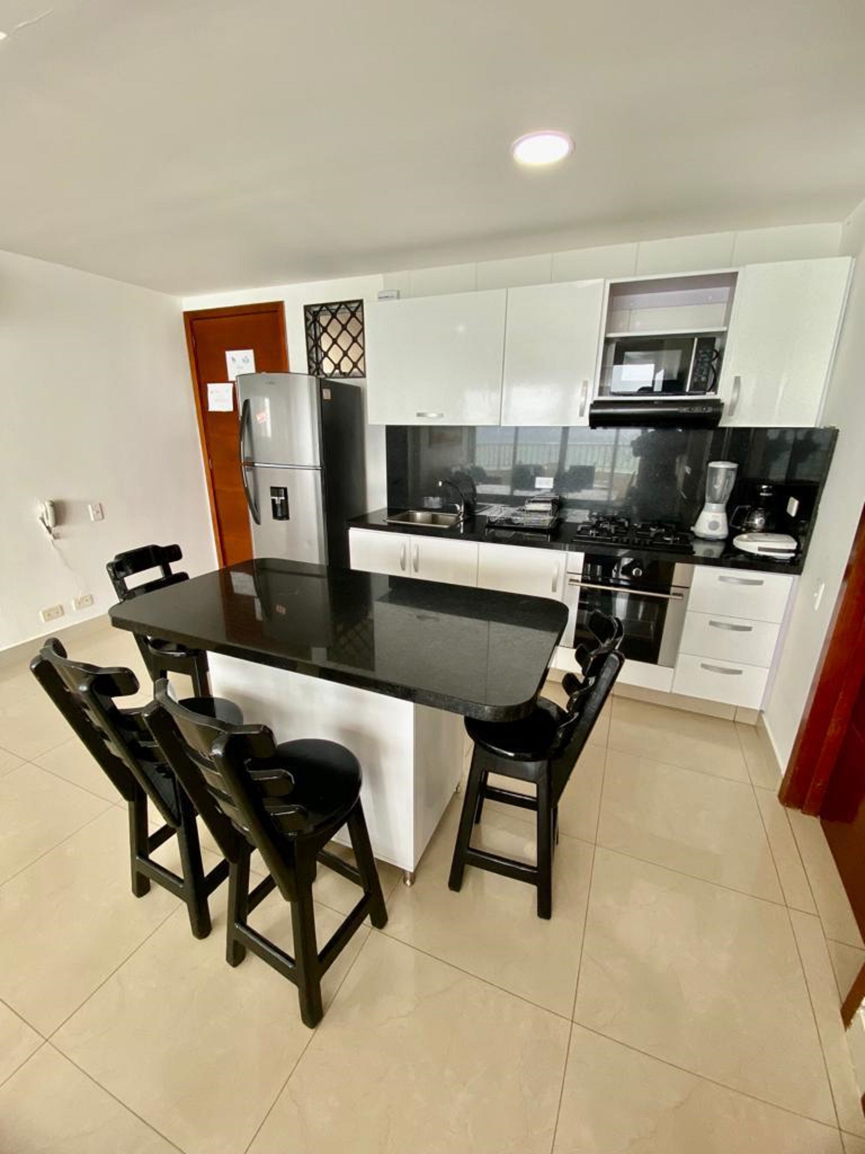 Cozinha moderna com bancada preta, armários brancos, micro-ondas embutido e fogão. Quatro cadeiras pretas estão dispostas ao redor de uma ilha central.