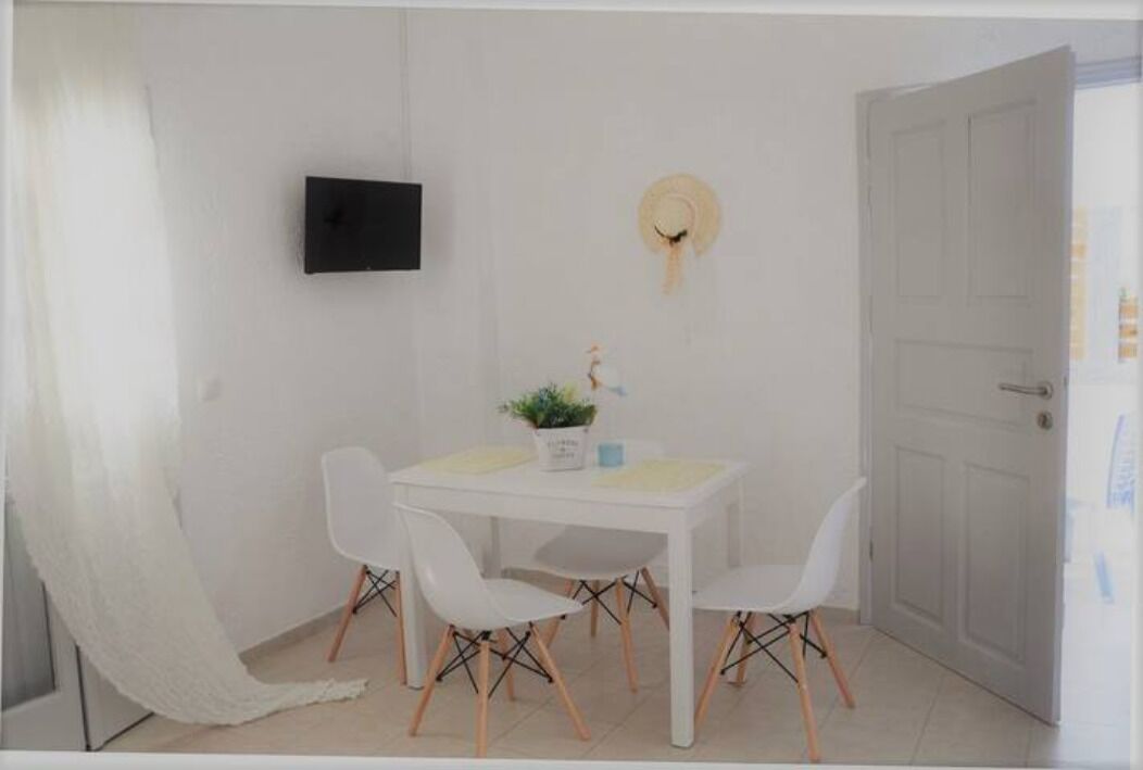 Uma pequena área de refeições branca, com uma mesa e quatro cadeiras, uma televisão na parede e uma porta à direita.