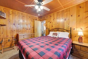 Quarto com painéis de madeira, uma cama, uma mesinha de cabeceira, um abajur e um ventilador de teto.