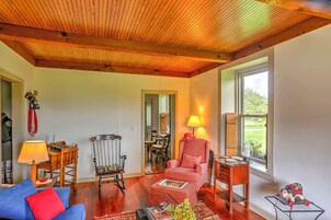 Uma sala de estar acolhedora com teto de madeira, uma cadeira de balanço, um poltrona vermelha xadrez, uma mesa com abajur e uma janela com vista para o verde.
