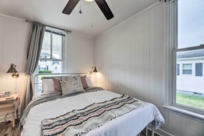 Quarto com cama, mesas de cabeceira, escrivaninha, janela com cortinas e ventilador de teto.
