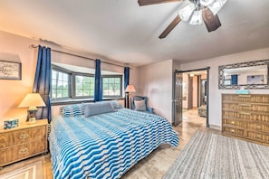 Quarto com uma cama grande, mesa de cabeceira de madeira, cômoda, ventilador de teto e janela com cortinas.