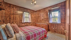 Quarto com painéis de madeira, colcha xadrez e duas janelas com cortinas.