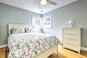 Quarto com cama, penteadeira, mesa de cabeceira, ventilador de teto e janela com vista para árvores.