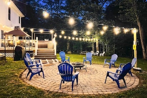 Uma área de pátio com cadeiras Adirondack azuis dispostas ao redor de um braseiro central, iluminada por luzes de fada.