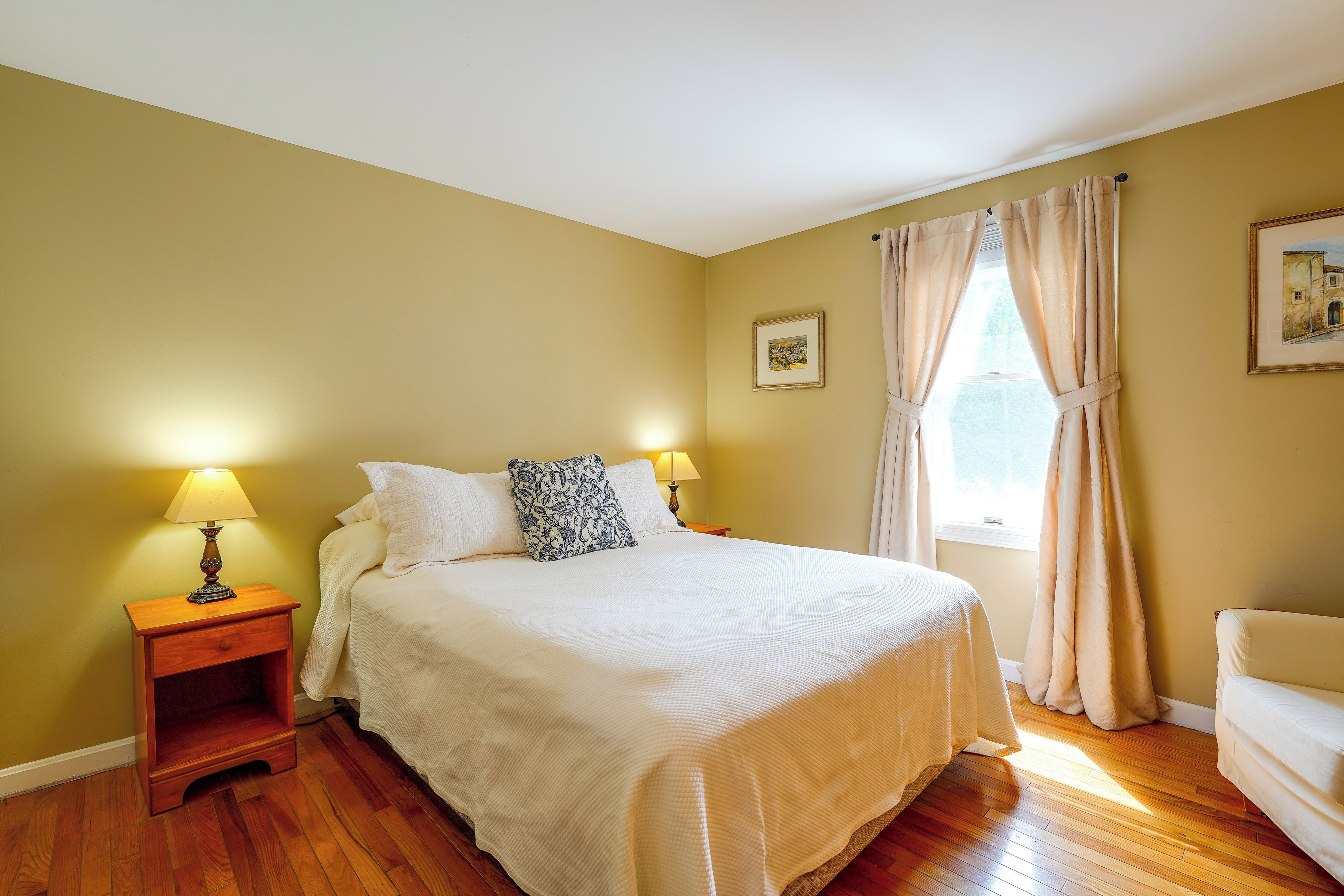 Quarto com cama, mesas de cabeceira, abajur, janela com cortinas e um quadro na parede.
