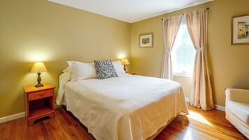 Quarto com cama, mesas de cabeceira, abajur, janela com cortinas e um quadro na parede.