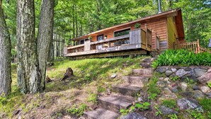 Uma cabana com fachada de madeira, construída sobre um alicerce de pedra, rodeada por árvores e uma escadaria de pedra.