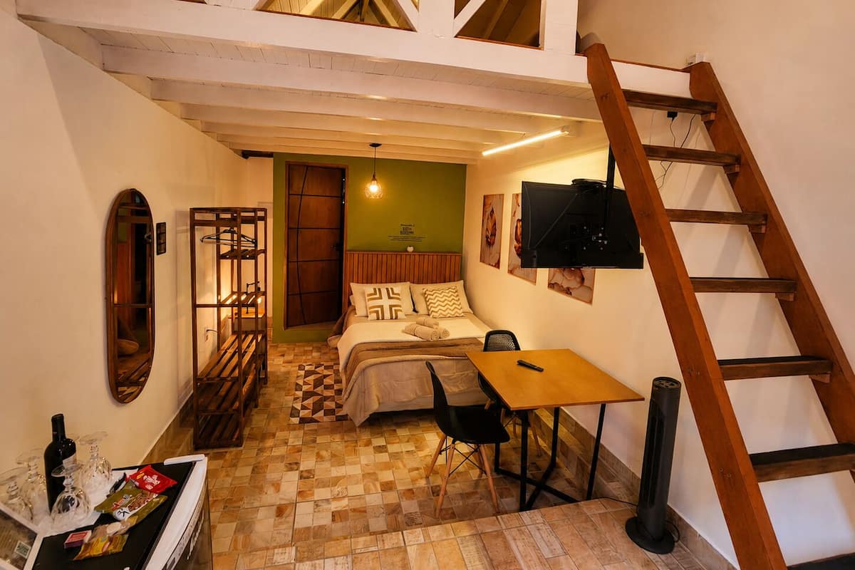 Quarto compacto com área de dormir no mezanino, escada de madeira, cama com almofadas estampadas, mesa pequena e TV fixada na parede.