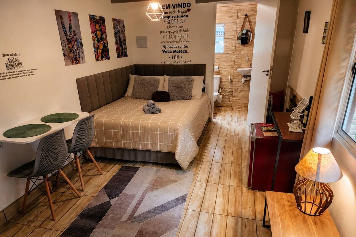Quarto com cama, escrivaninha, cadeiras, tapete e obras de arte nas paredes.
