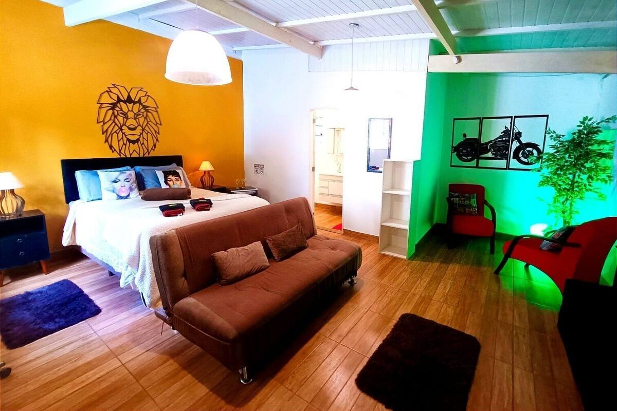 Quarto de hotel moderno com cama, sofá, área de jantar e parede verde com arte de motocicletas.
